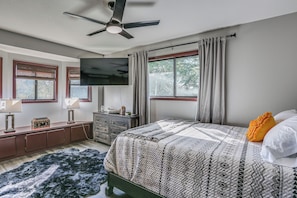 Quarto com cama, ventoinha no teto, televisão, penteadeira e janela com cortinas.