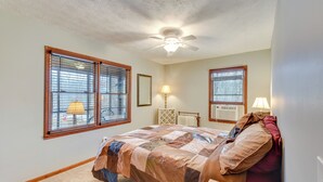 Quarto com cama, duas janelas com persianas, ventilador de teto e mesa de cabeceira com abajur.