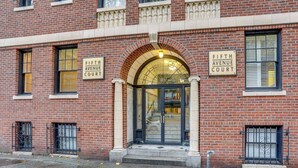 Um edifício de tijolos com uma grande entrada em arco, duas placas com a inscrição 'Fifth Avenue Court' e um conjunto de degraus que levam à entrada.