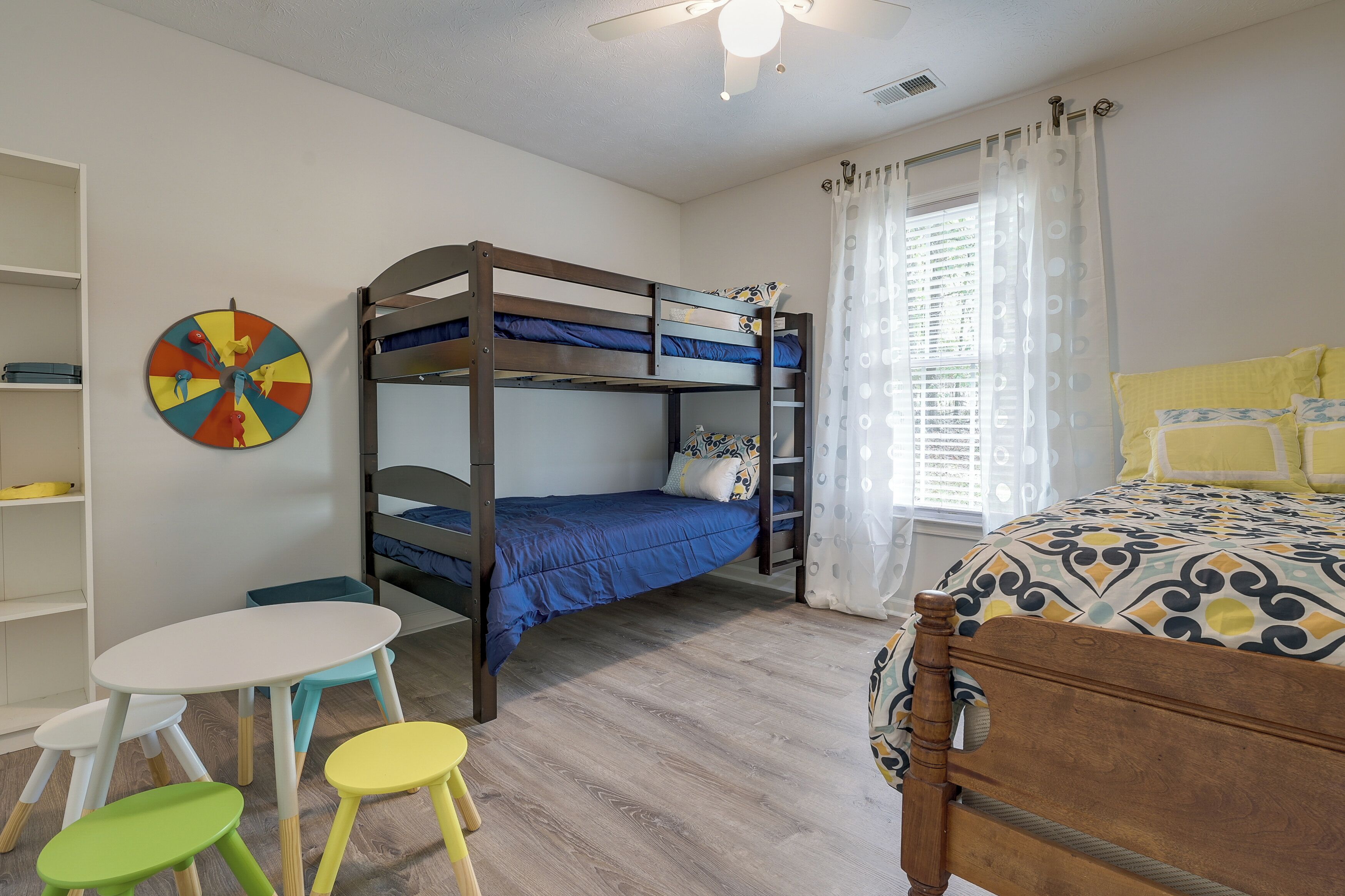 Quarto com beliche, ventilador de teto, relógio redondo na parede, mesa pequena com bancos e cama com roupa de cama estampada.