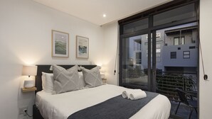 Uma cama bem arrumada com um cobertor escuro, duas luminárias fixadas na parede e duas obras de arte emolduradas acima.
