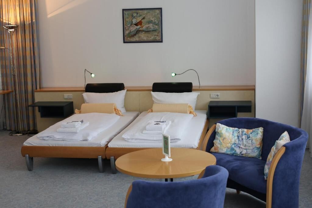 Quarto de hotel com duas camas de solteiro, uma mesa redonda de madeira, uma poltrona azul e um quadro na parede.