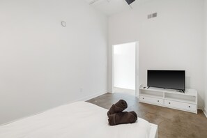 Quarto minimalista com uma cama, uma televisão sobre um suporte branco e uma porta que dá para outro espaço.