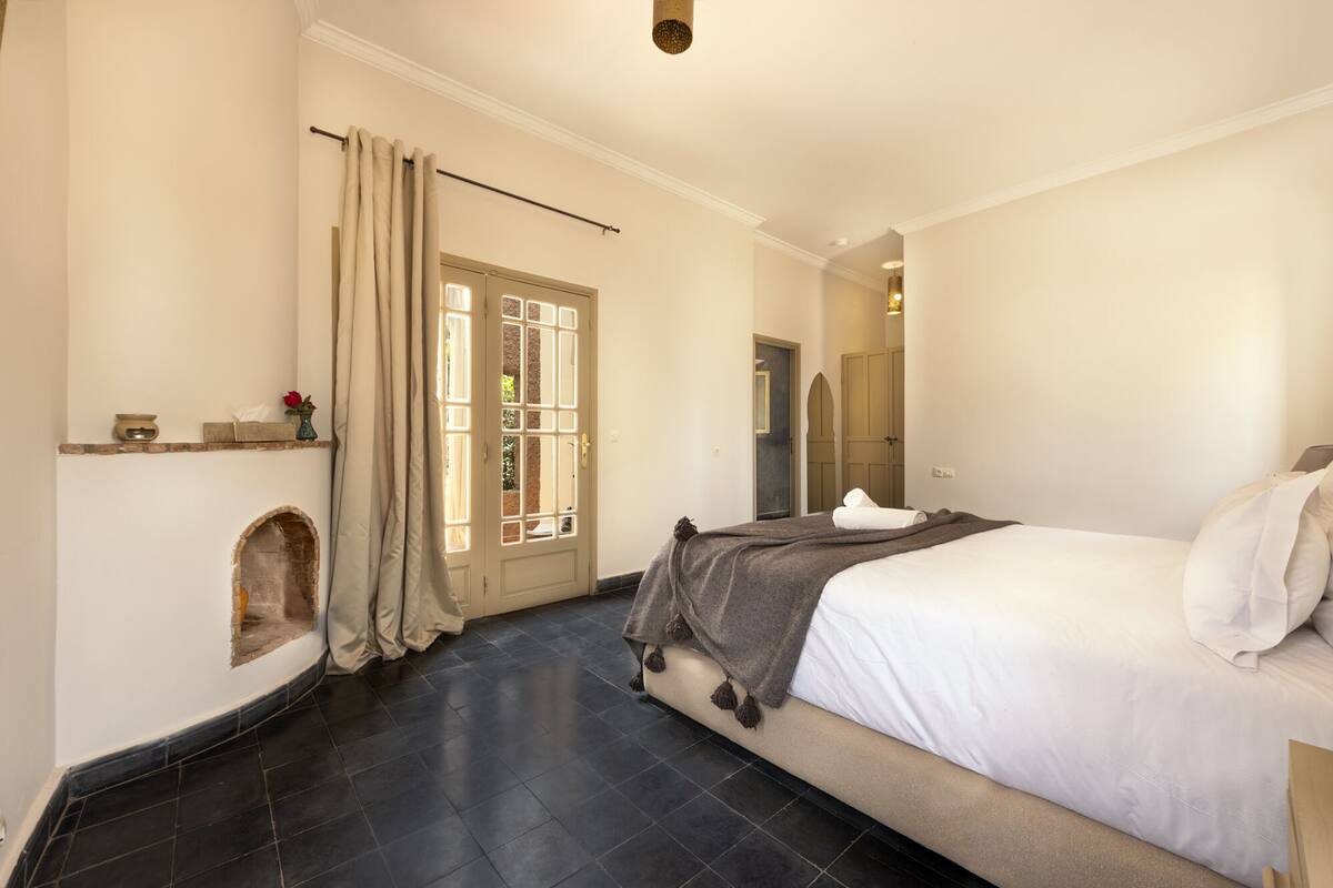 Quarto com cama, lareira, porta com painéis de vidro e uma pequena estante com objetos.