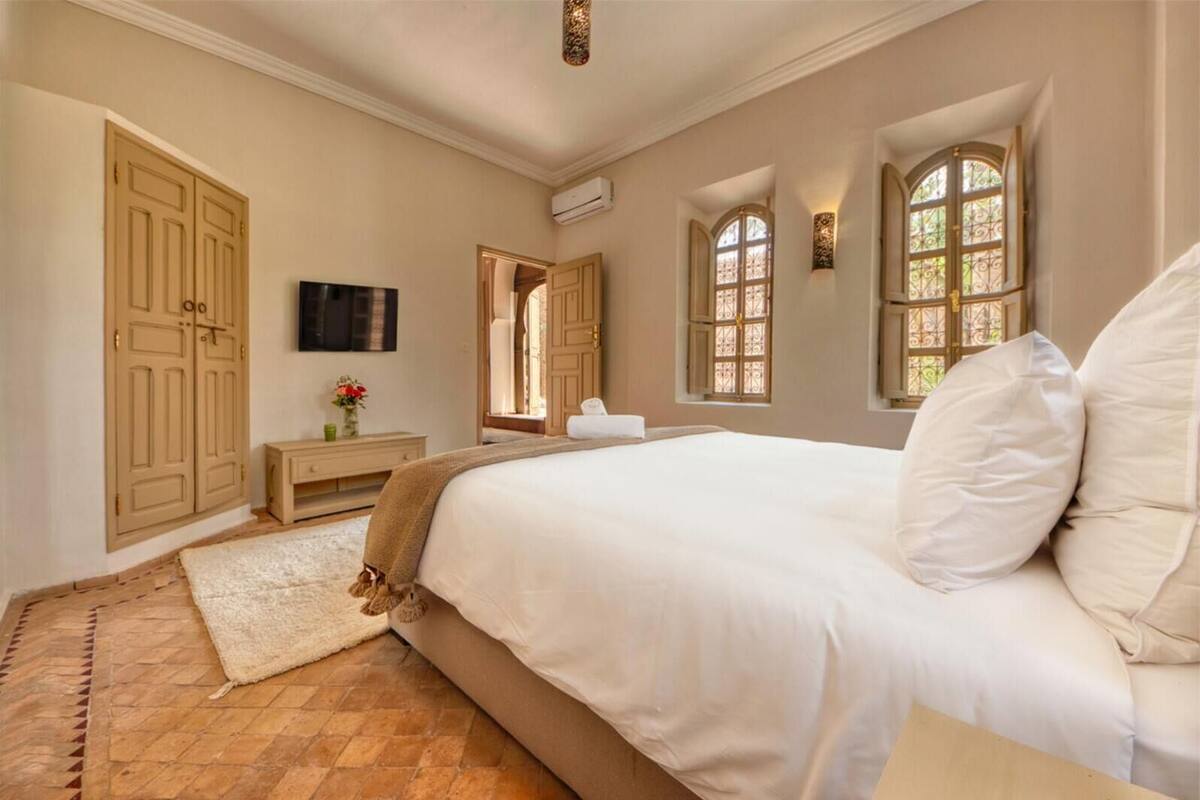 Quarto com cama grande, penteadeira, televisão e duas janelas com grades decorativas.