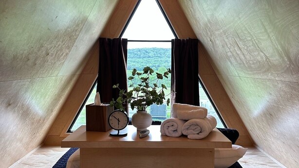 Um quarto acolhedor em formato triangular, com uma cama, uma mesinha de cabeceira com um relógio, um vaso com uma planta e toalhas enroladas.