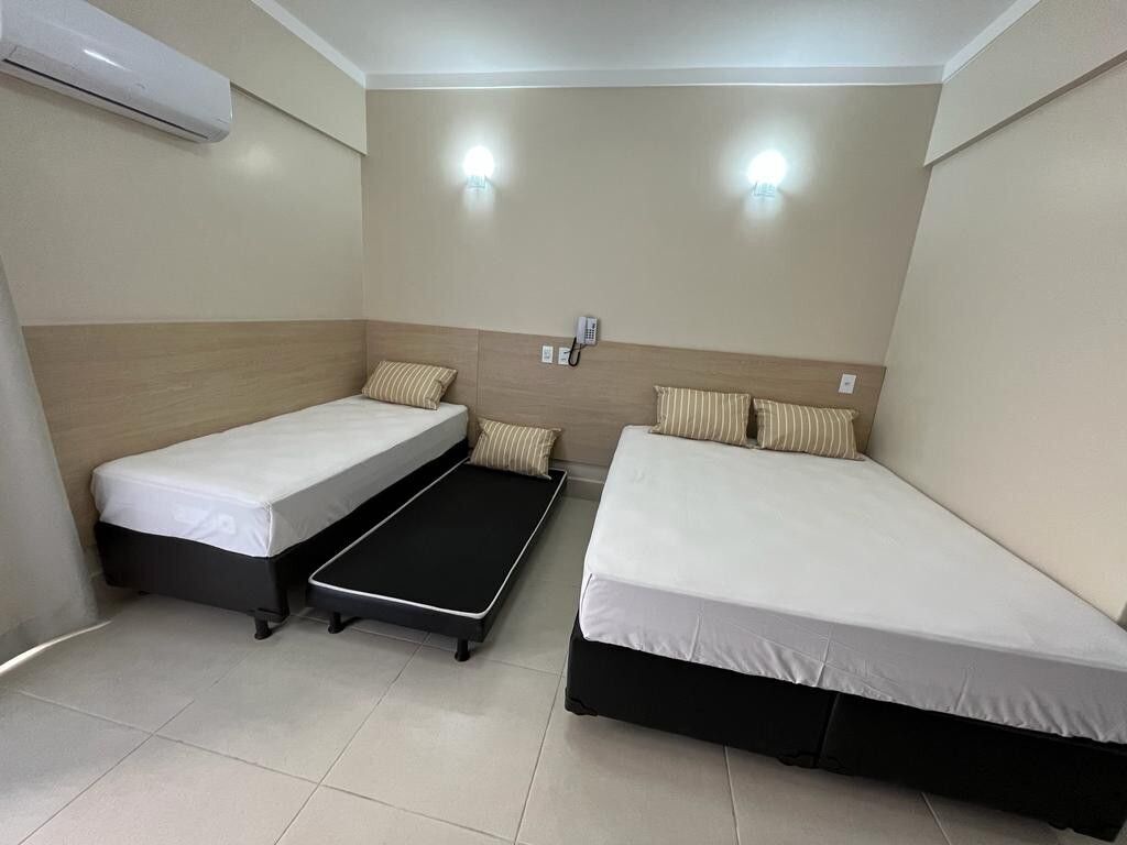 Quarto de hotel com duas camas de solteiro, uma pequena mesa, aparelho de ar condicionado e duas luminárias fixadas na parede.