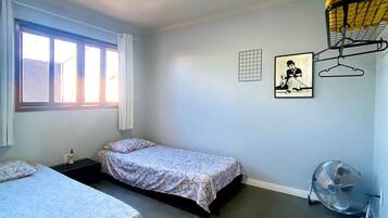 Quarto pequeno e pouco mobiliado, com duas camas de solteiro, ventoinha no teto, janela com cortinas e um varal fixado na parede.