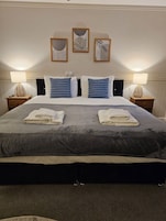 Cama bem arrumada com duas toalhas dobradas, um edredom cinza escuro e um travesseiro branco. Duas luminárias de mesa com bases decorativas, uma obra de arte na parede acima da cama e uma mesa de cabeceira de madeira com um controle remoto.