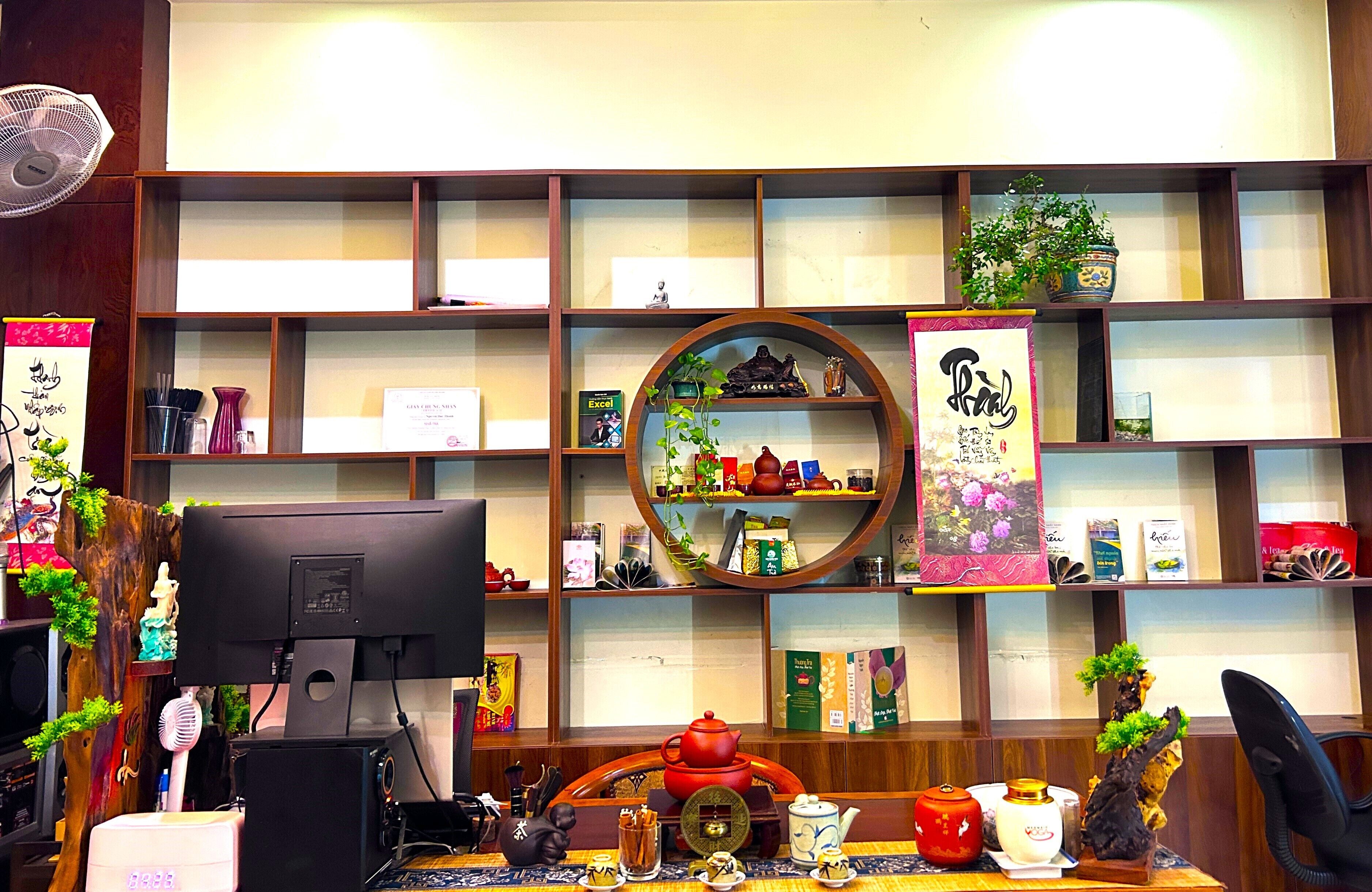 Escritório bem organizado com computador, estantes com livros e objetos decorativos, mesa com bule e ventilador.