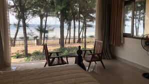 Quarto com uma janela grande de frente para a praia, uma cama, uma cadeira e um ventilador.