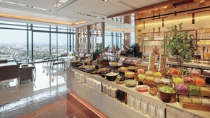 Buffet com diversas opções de comida, área de estar com cadeiras e janelas amplas com vista para a cidade.