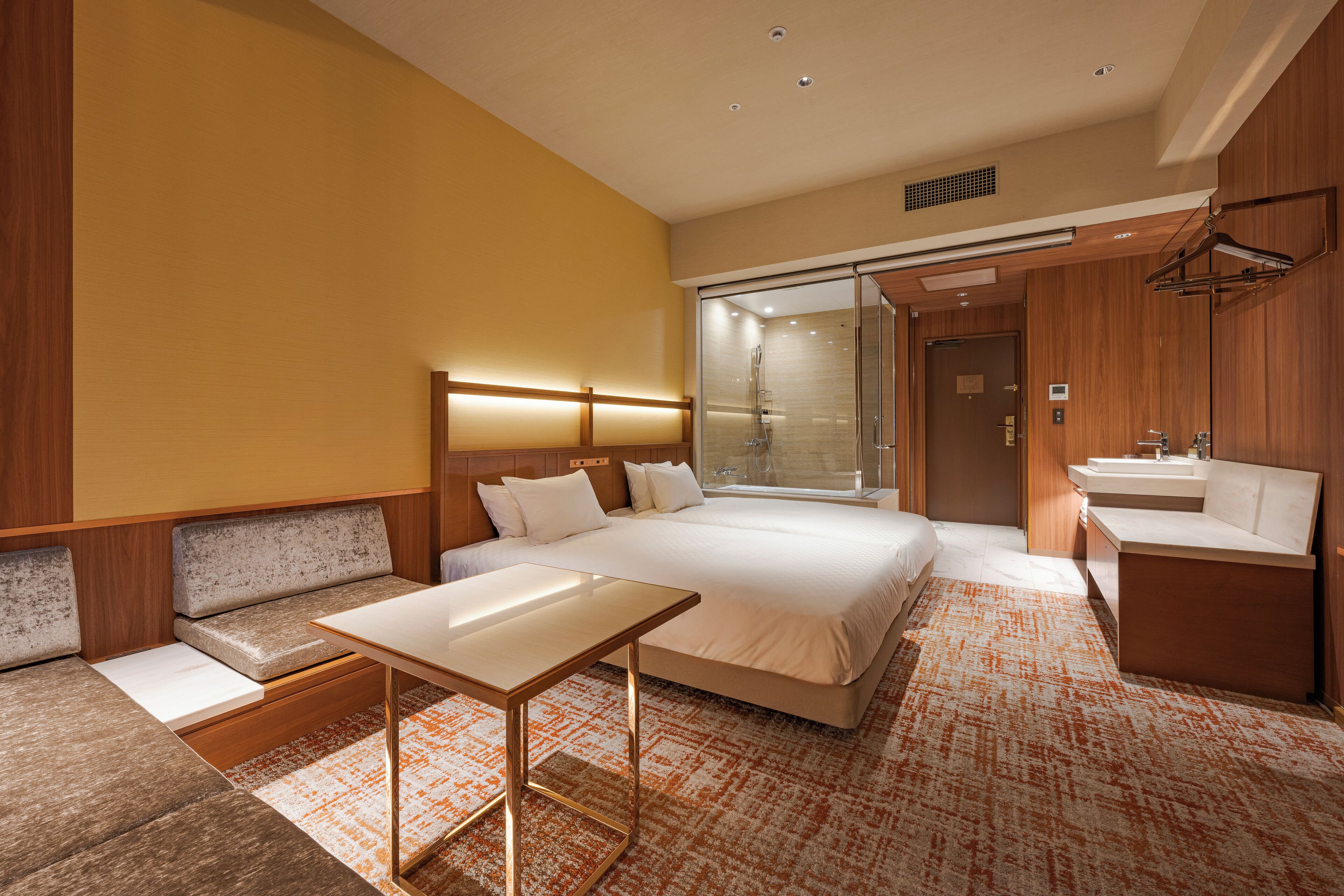 Quarto de hotel com uma cama grande, uma mesa de cabeceira, um espaço de estar com um banco, área de chuveiro e banheiro com pia e espelho.