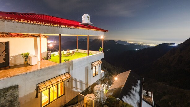 Um edifício de dois andares com terraço no telhado, janelas amplas e telhado vermelho.