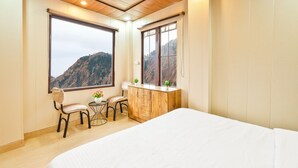 Quarto com uma cama grande, guarda-roupa de madeira, duas cadeiras, uma mesa pequena e duas janelas com vista para as montanhas.
