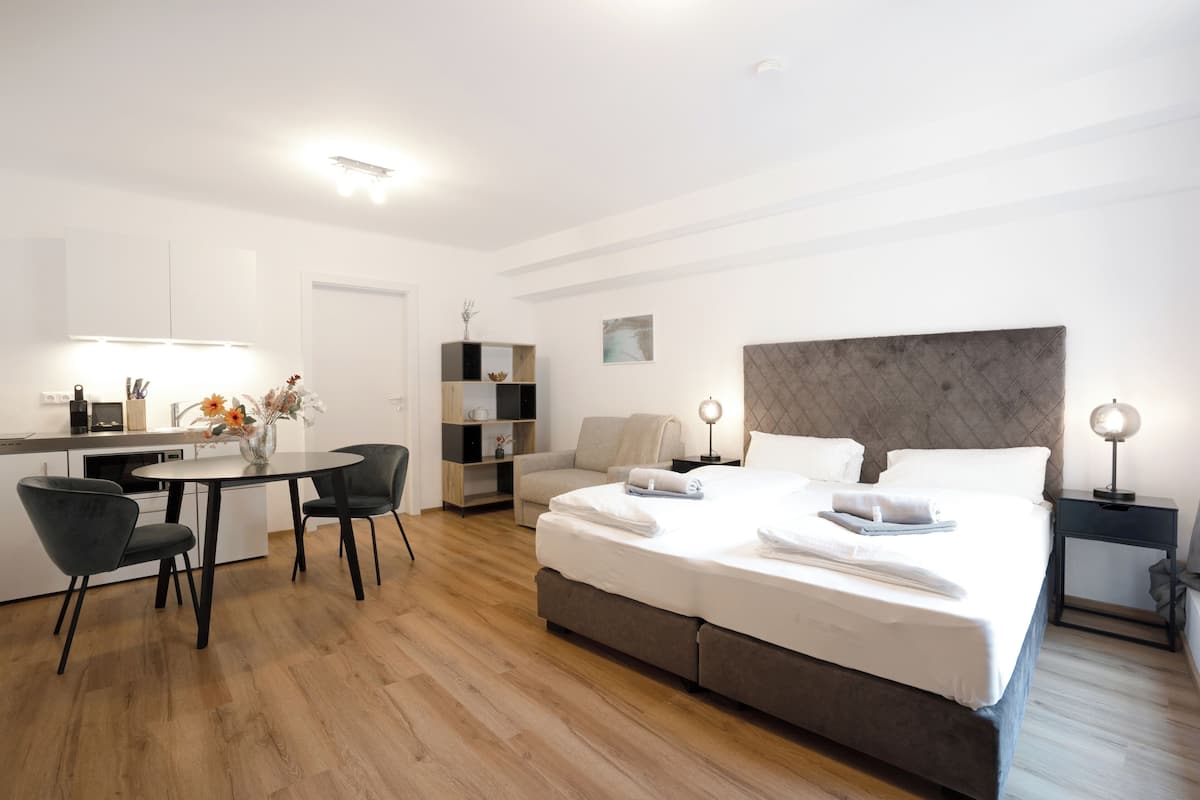 Quarto de hotel com cama, mesas de cabeceira, escrivaninha com cadeiras, estante de livros e um pequeno espaço de estar.