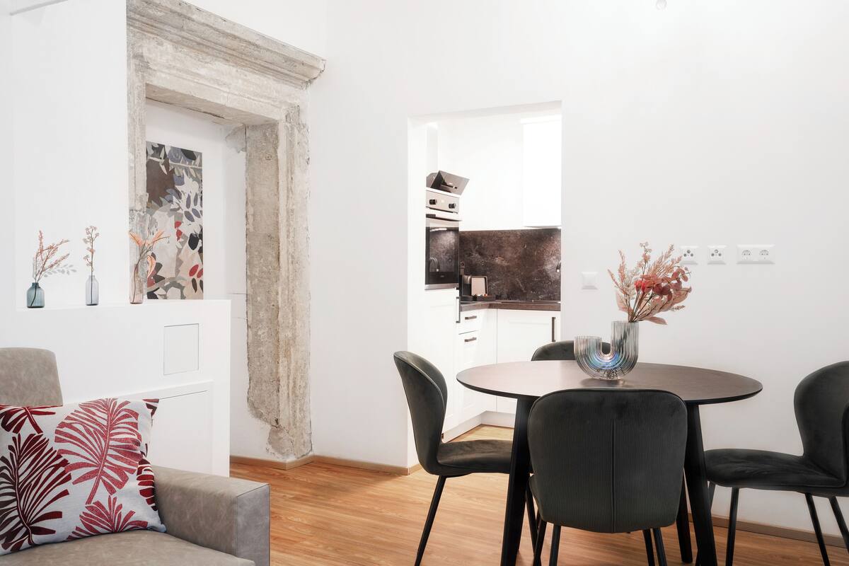 Uma sala de estar moderna com uma mesa de jantar redonda, cadeiras escuras e um sofá cinza com almofada estampada.