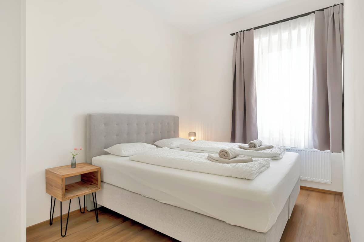 Cama bem arrumada com lençóis brancos e toalhas dobradas, mesa de cabeceira com um vaso, e janela com cortinas.