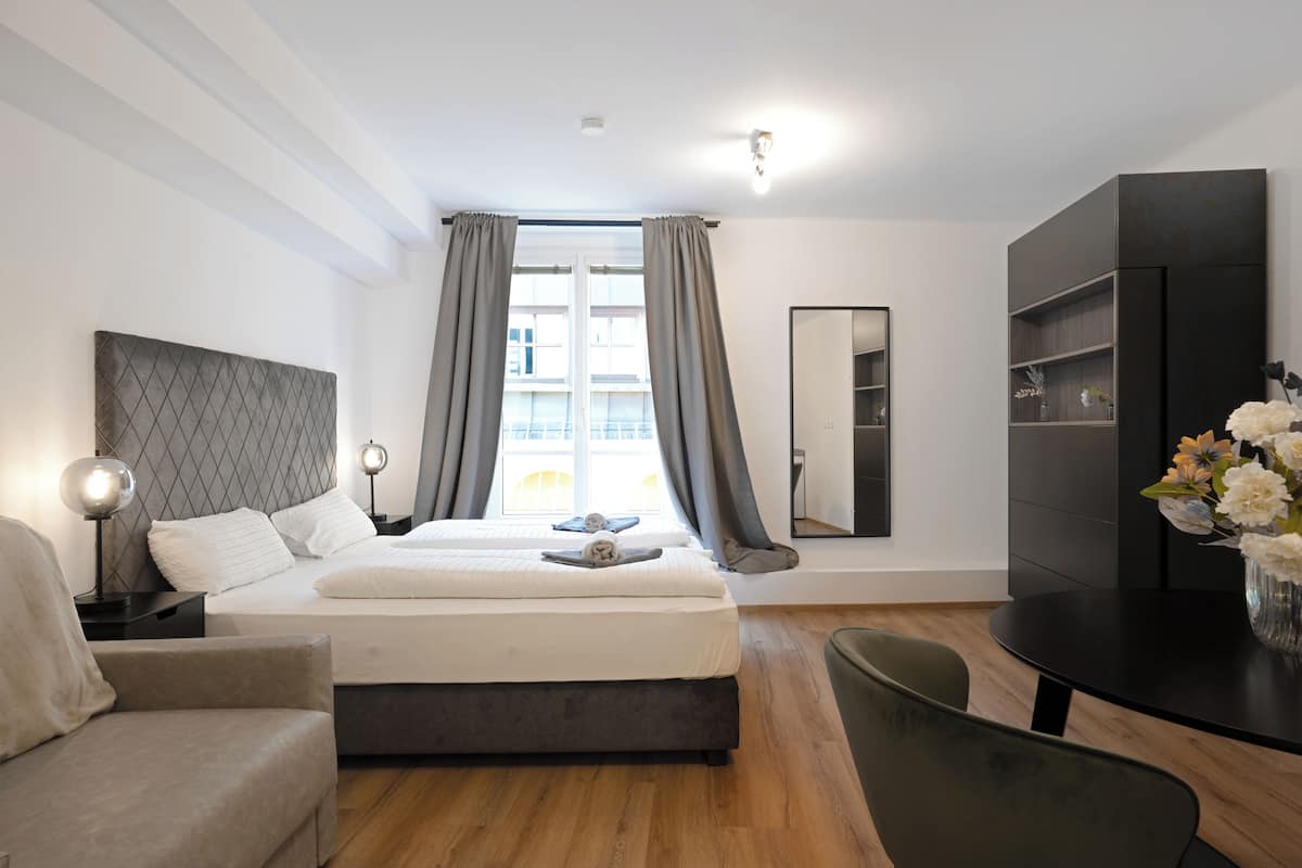Quarto de hotel moderno com cama, mesas de cabeceira, escrivaninha, cadeira, espelho e televisão.