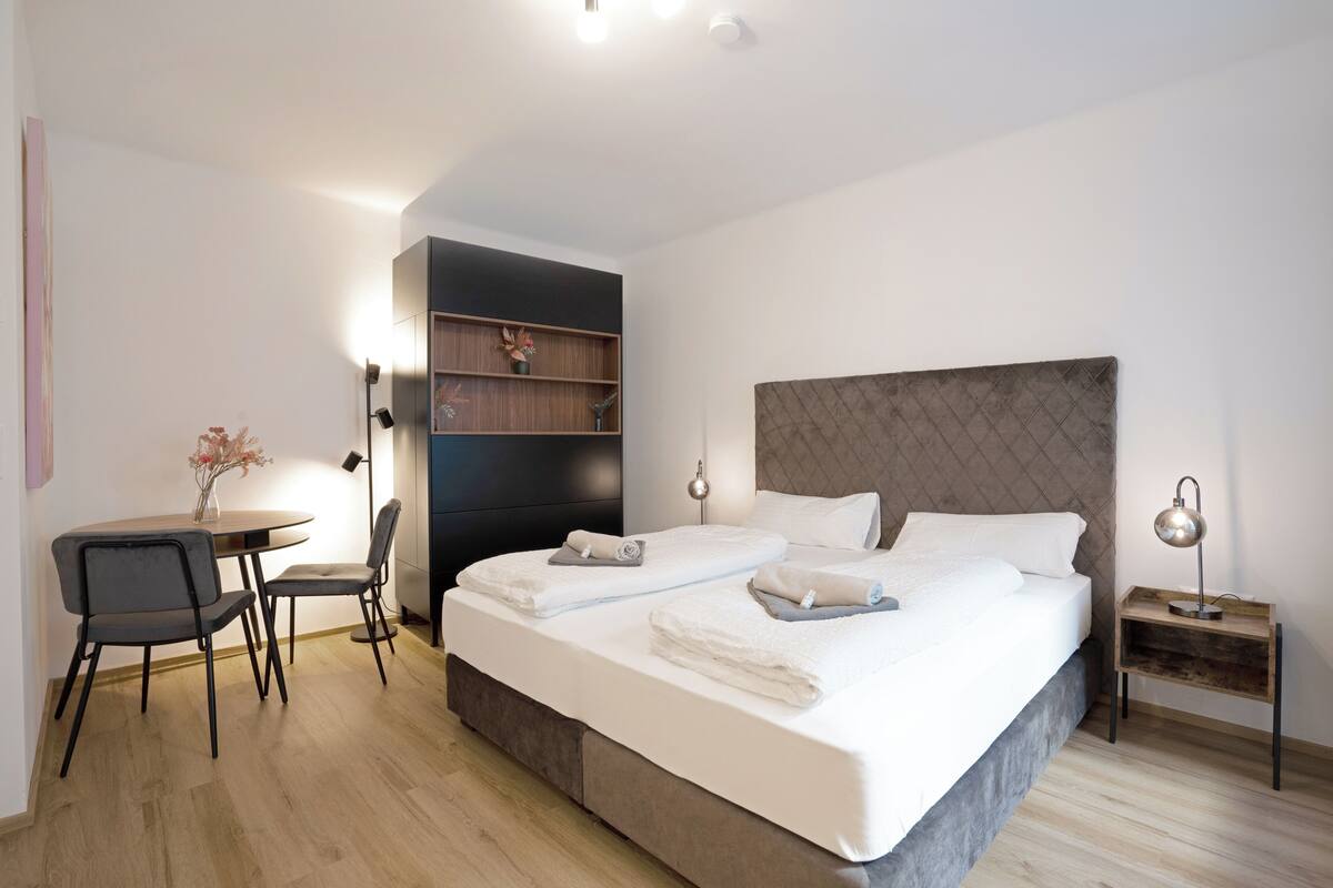 Quarto de hotel moderno com uma cama grande, uma escrivaninha com duas cadeiras, uma mesinha com um vaso e uma mesa de cabeceira com uma luminária.