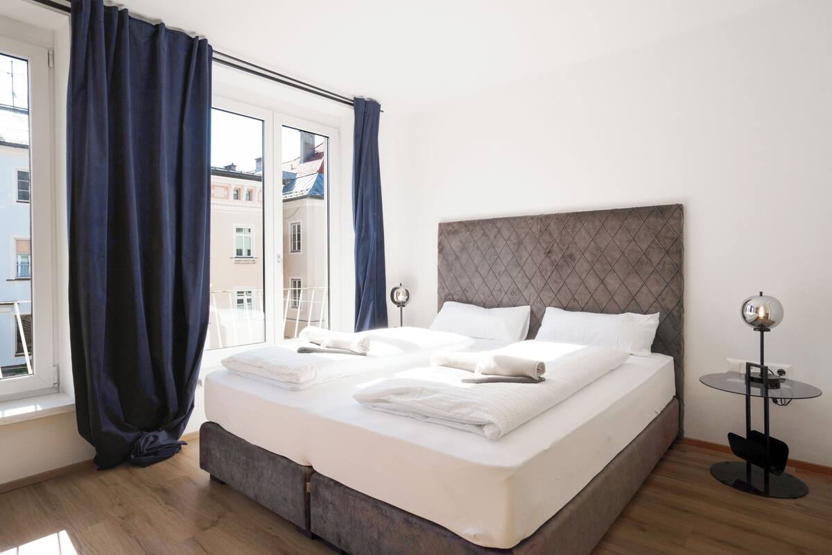 Uma cama bem arrumada com lençóis brancos, cabeceira cinza e mesa de cabeceira com abajur.