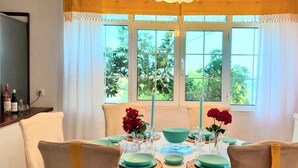 Uma sala de jantar com uma mesa posta para quatro pessoas, janelas grandes com cortinas e vista para o verde lá fora.