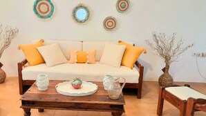 Uma sala de estar aconchegante com um sofá branco, mesa de centro de madeira e duas poltronas. Há elementos decorativos nas paredes acima do sofá e um vaso com galhos no chão.