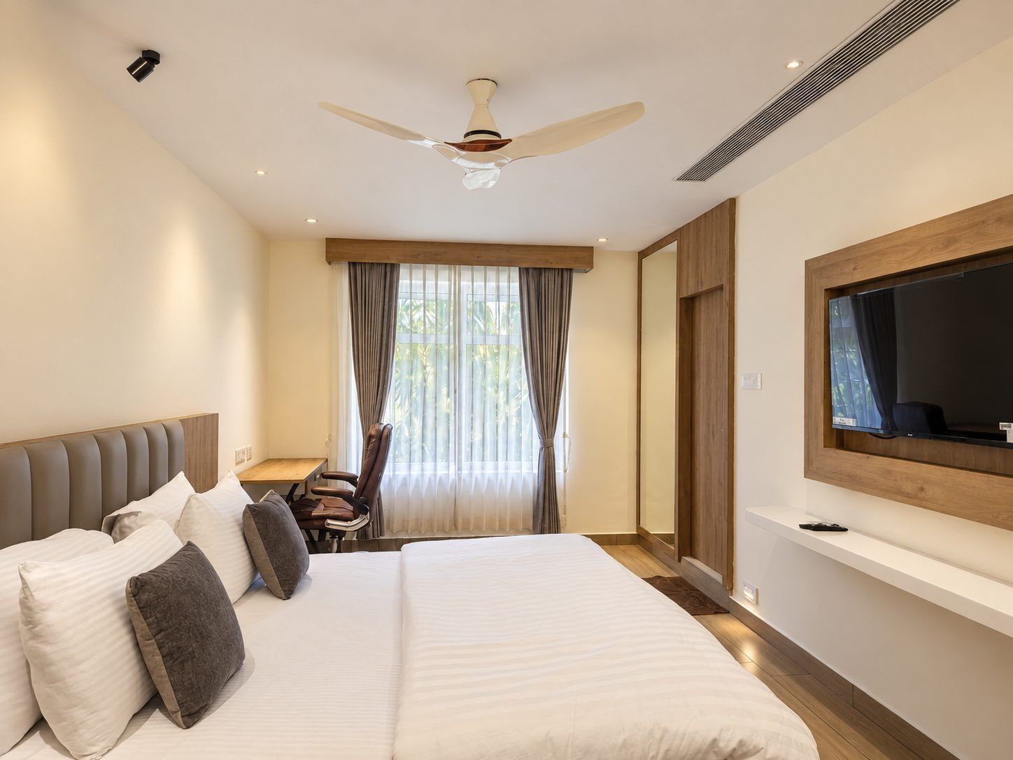 Quarto de hotel com uma cama grande, uma escrivaninha, uma cadeira, um ventilador de teto e uma TV fixada na parede.