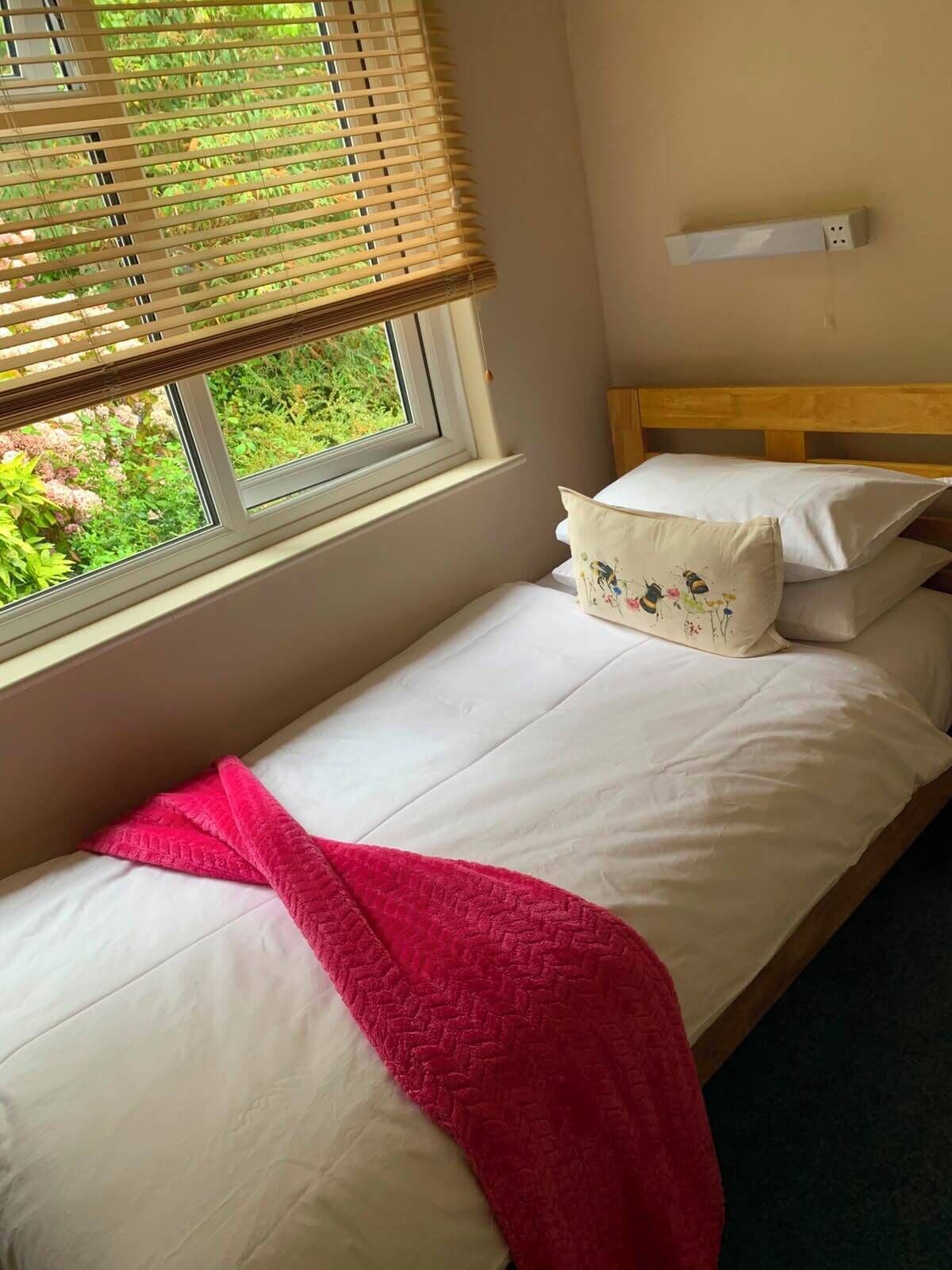 Uma cama de solteiro com roupa de cama branca, um cobertor rosa e um travesseiro com padrão floral. Há uma janela com persianas e um luminário fixado na parede sobre a cama.