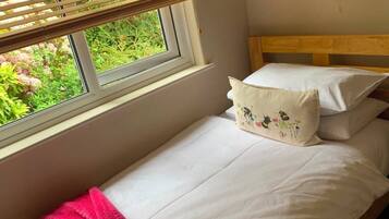 Uma cama de solteiro com roupa de cama branca, um cobertor rosa e um travesseiro com padrão floral. Há uma janela com persianas e um luminário fixado na parede sobre a cama.