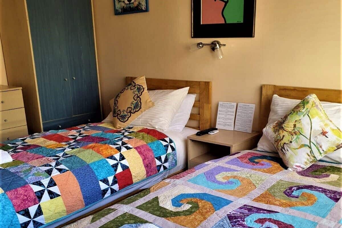 Quarto de hotel com duas camas, cada uma com um edredom colorido, uma cabeceira de madeira, uma mesa de cabeceira com abajur e um quadro na parede.