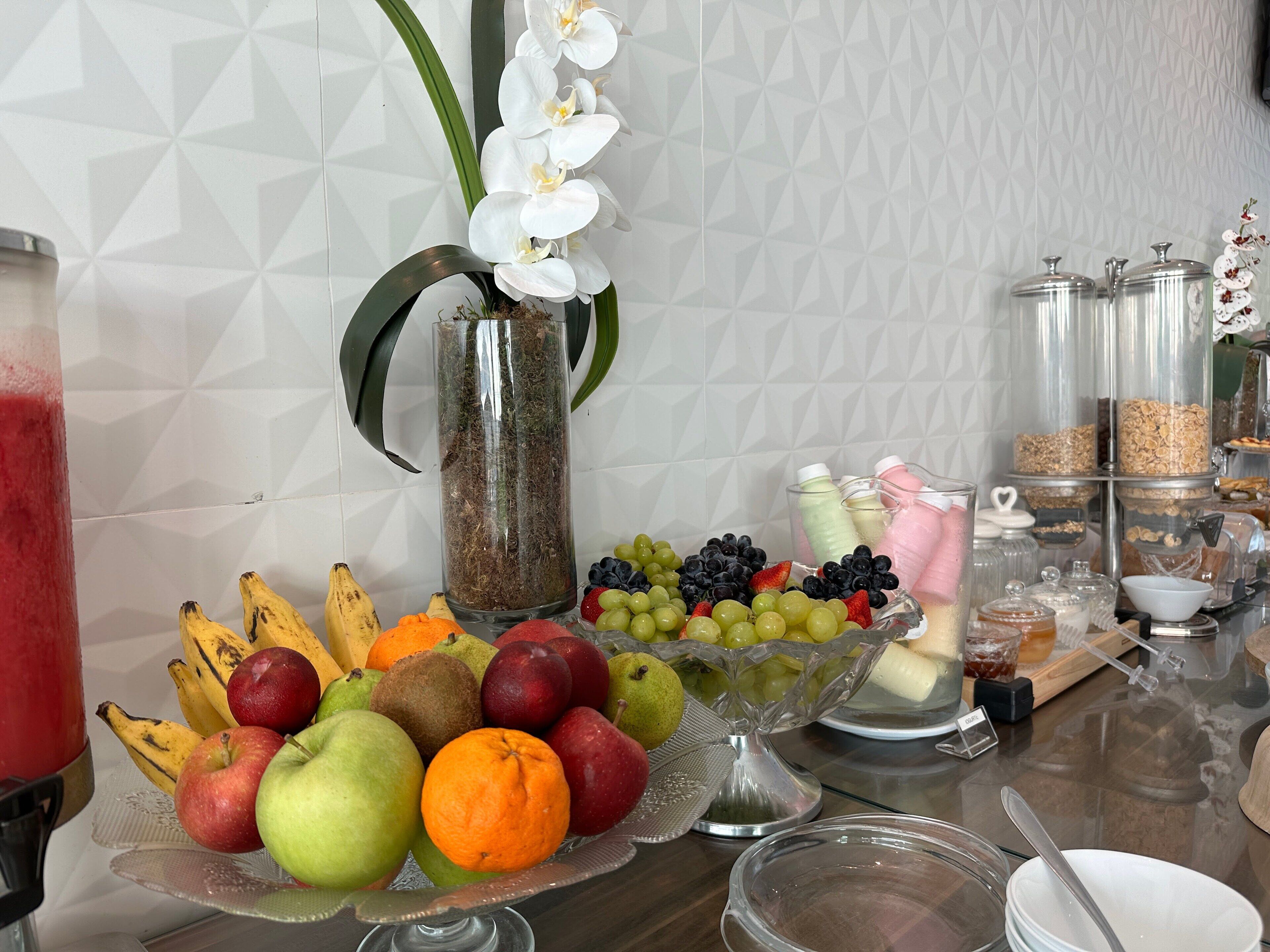Um buffet de café da manhã com diversas frutas, um vaso com flores brancas e recipientes de comida.