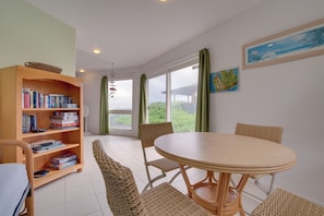 Uma área de refeições com uma mesa redonda e cadeiras de vime, uma estante com livros e vista para o verde através de amplas janelas.