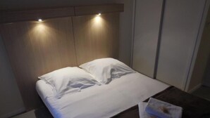 Cama bem arrumada com lençóis brancos e um cobertor marrom escuro, com uma toalha dobrada ao lado.