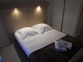 Cama bem arrumada com lençóis brancos e um cobertor marrom escuro, com uma toalha dobrada ao lado.