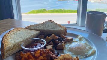 Prato de café da manhã com torradas, ovos, bacon e batatas, com vista para o mar pela janela.