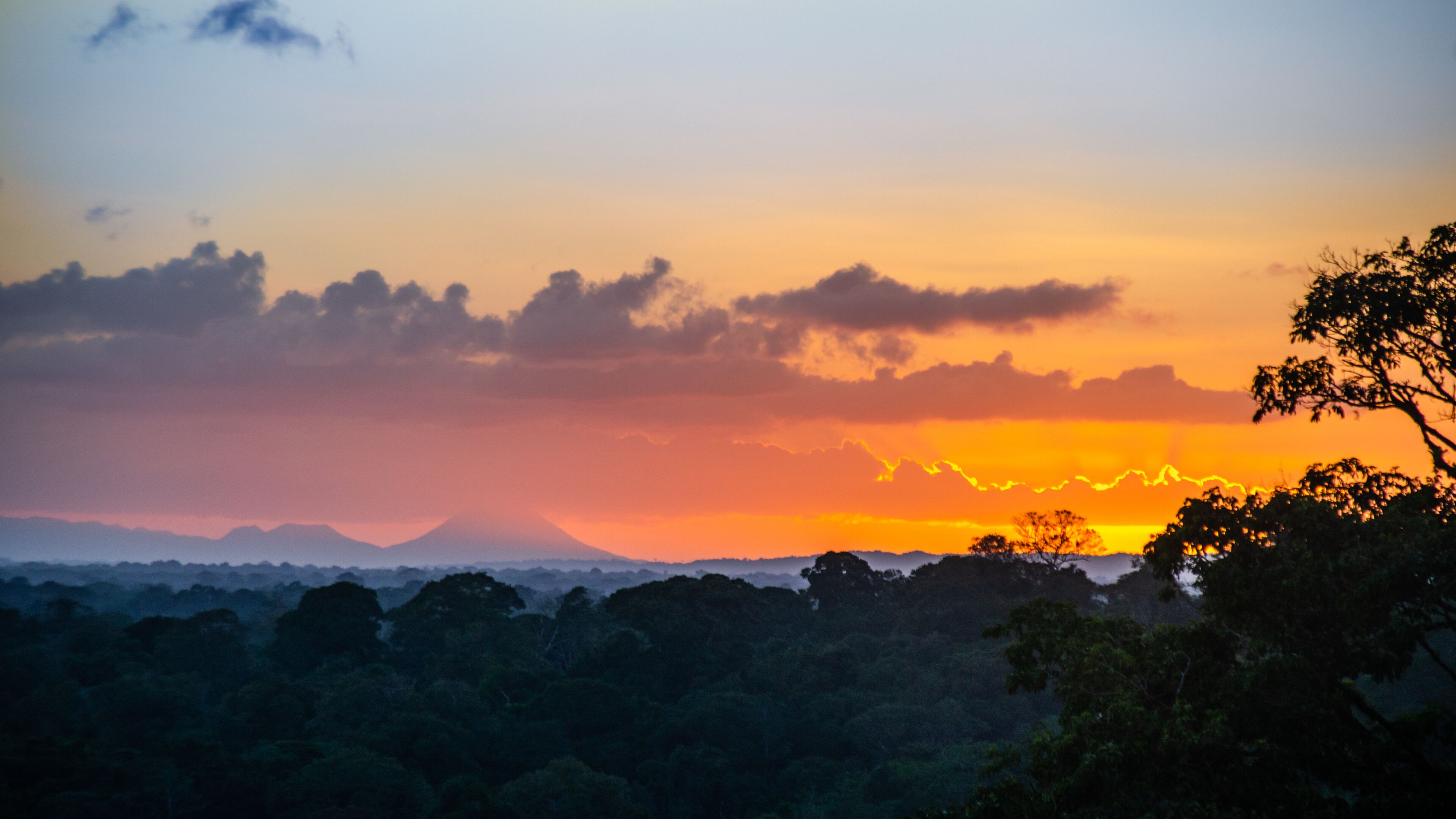 Paisagem com pôr do sol sobre uma área arborizada e montanhas ao longe.