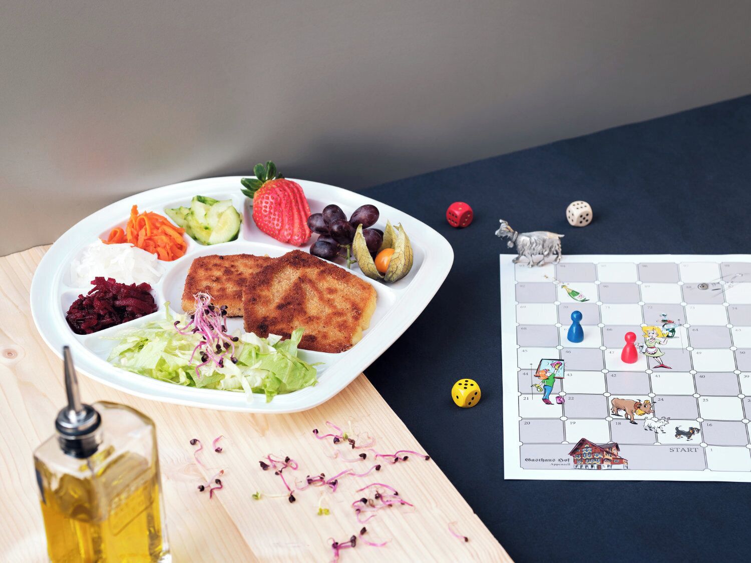Uma refeição com peixe empanado, legumes e frutas em um prato, um jogo de tabuleiro e uma garrafa de óleo sobre uma mesa.