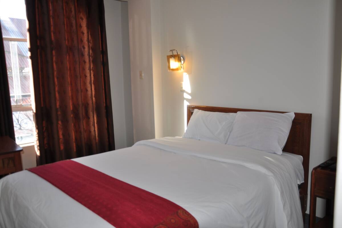 Uma cama bem arrumada com lençóis brancos e um cobertor vermelho, um luminária fixada na parede e uma janela com cortinas.