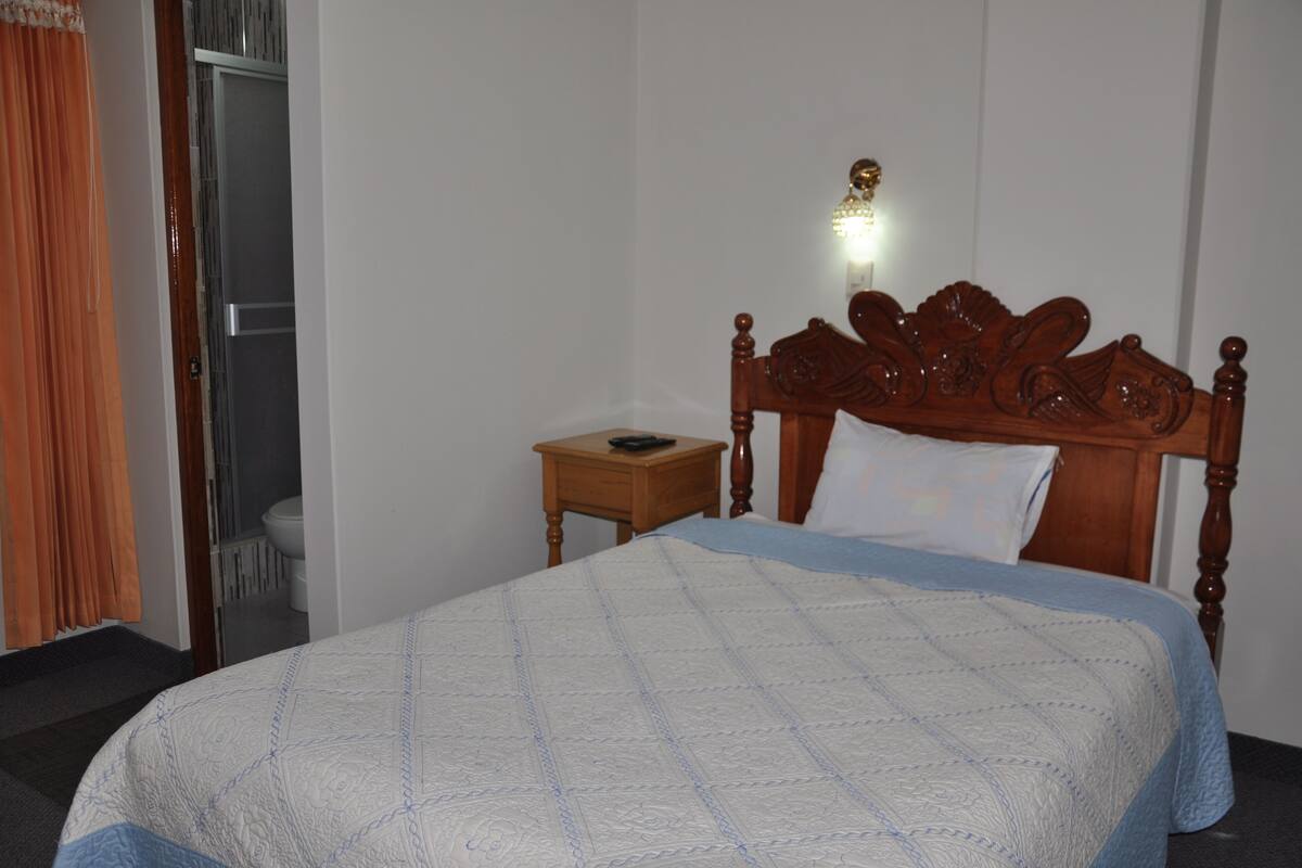 Uma cama bem arrumada com peseira de madeira, mesa de cabeceira com abajur, e um banheiro visível por uma porta aberta.