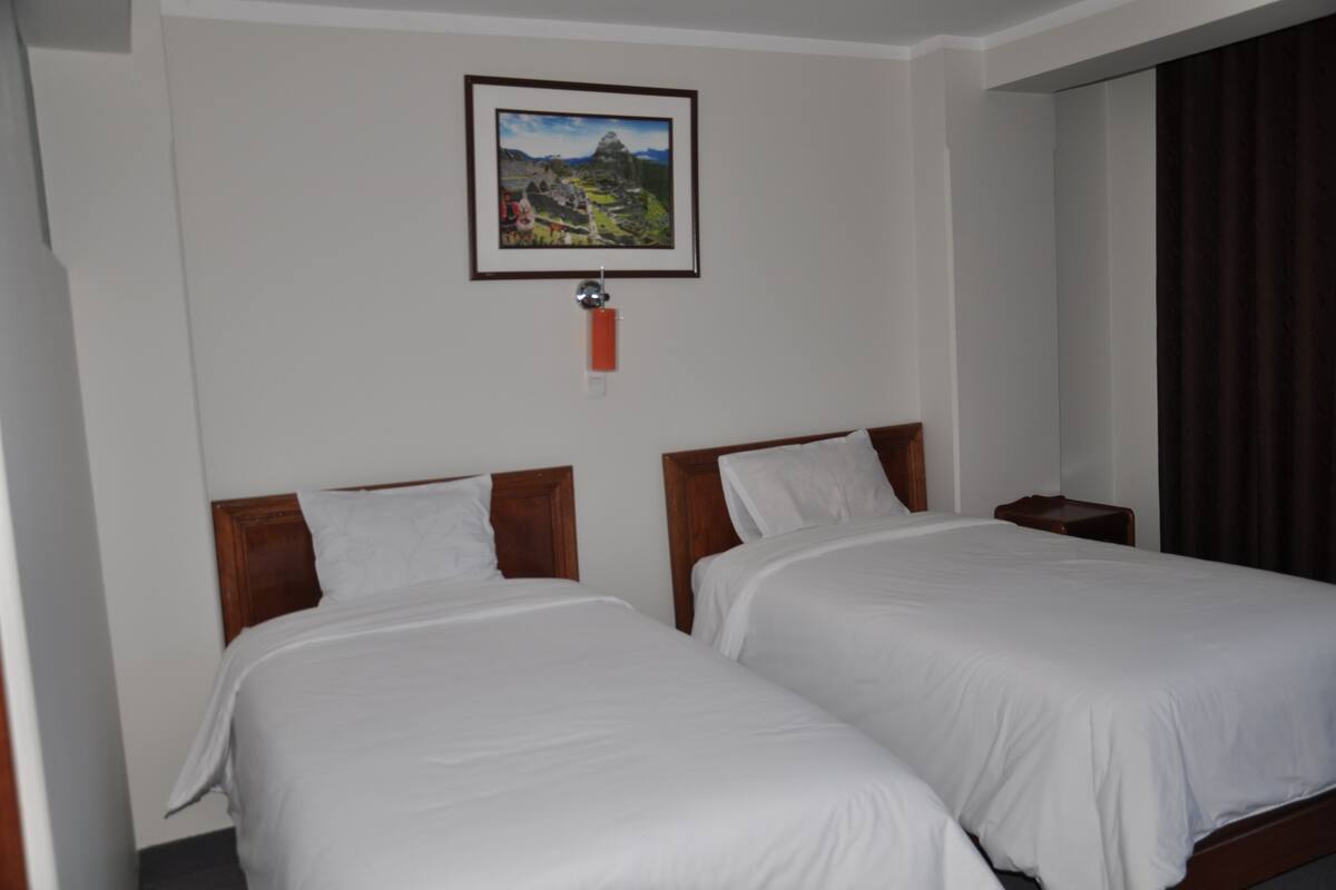 Quarto de hotel com duas camas de solteiro, cabeceira de madeira, quadro na parede e janela com cortinas.