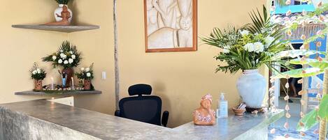 Uma área de recepção com balcão de mármore, uma cadeira de escritório preta, um vaso grande com flores e um quadro pendurado na parede.