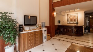 Uma área de recepção de hotel com balcão de madeira, TV de tela plana, dispensador de água e um painel decorativo na parede.