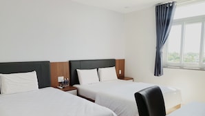 Quarto de hotel com duas camas de solteiro, uma mesa de cabeceira de madeira com um controle remoto e garrafas de água, uma escrivaninha e uma janela com vista para o verde.