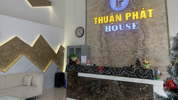 Uma área de recepção de hotel com design moderno, incluindo um balcão de atendimento, um relógio e uma placa fixada na parede com a inscrição "THUAN PHAT HOUSE".