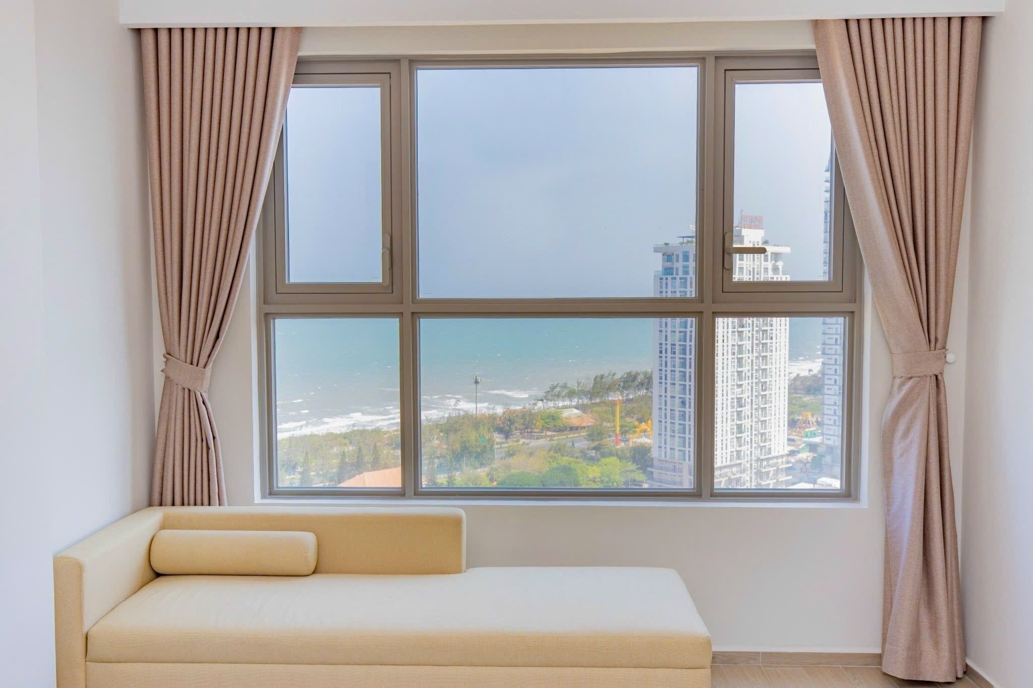 Quarto com uma janela grande, cortinas bege e um sofá bege.