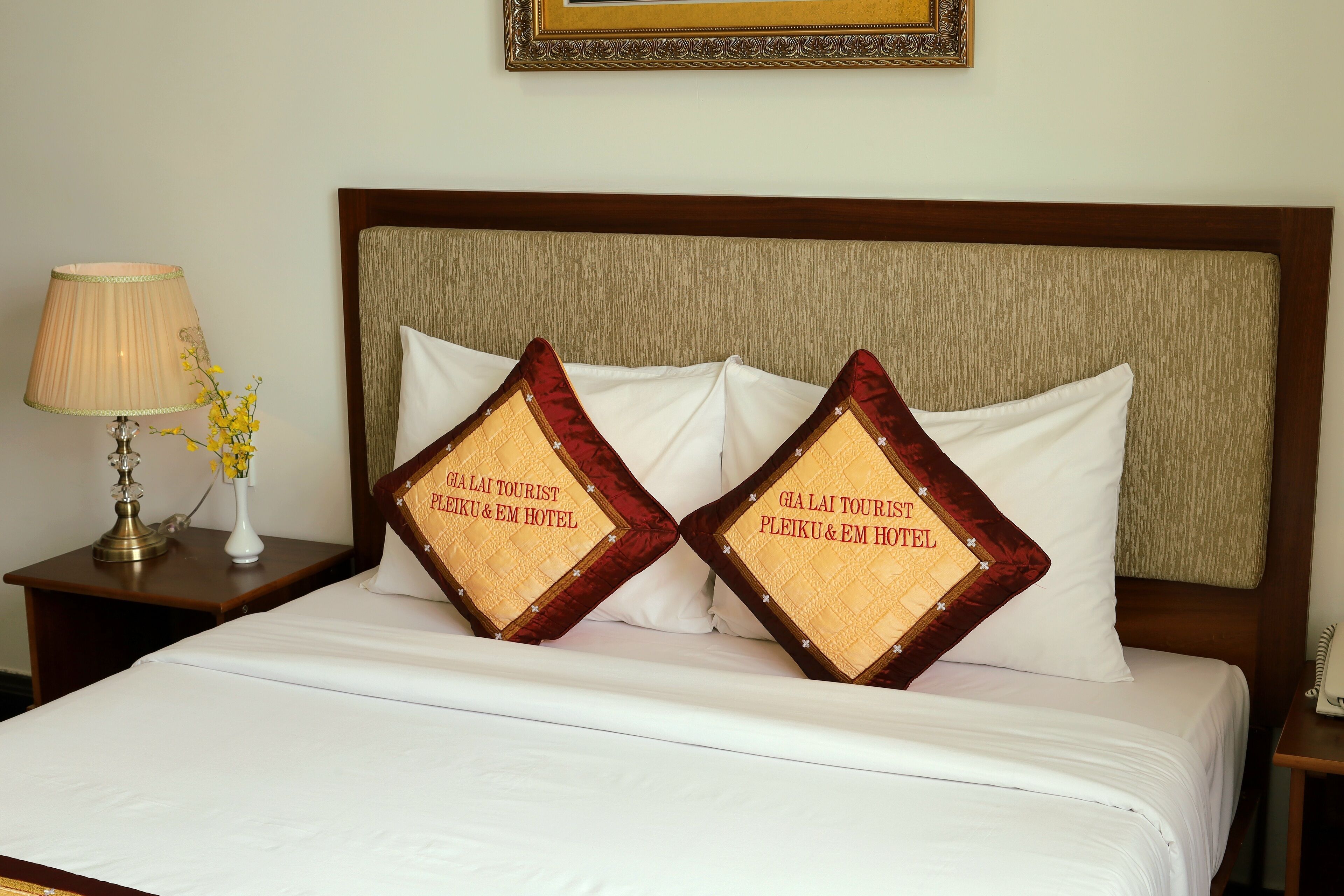 Uma cama bem arrumada com lençóis brancos, duas almofadas decorativas com texto, um abajur e um vaso com flores amarelas.