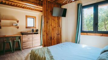 Quarto com teto de madeira, uma cama, televisão montada na parede, janela com cortinas e uma escrivaninha com cadeiras.