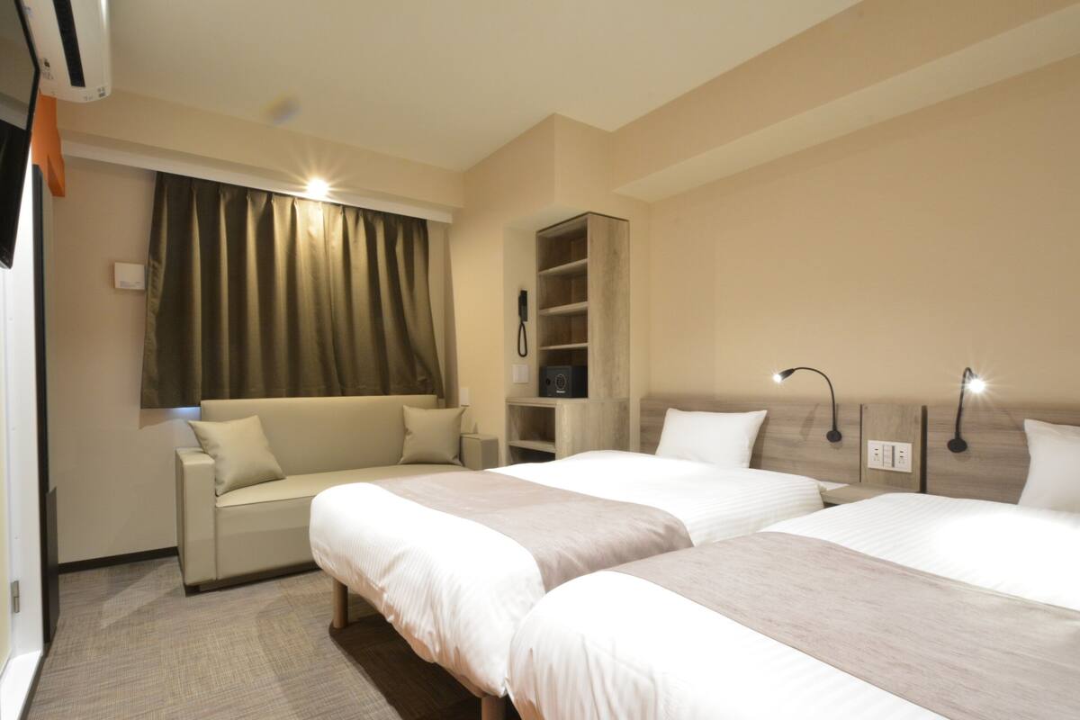 Quarto de hotel com duas camas, um sofá, uma escrivaninha e uma janela com cortinas.
