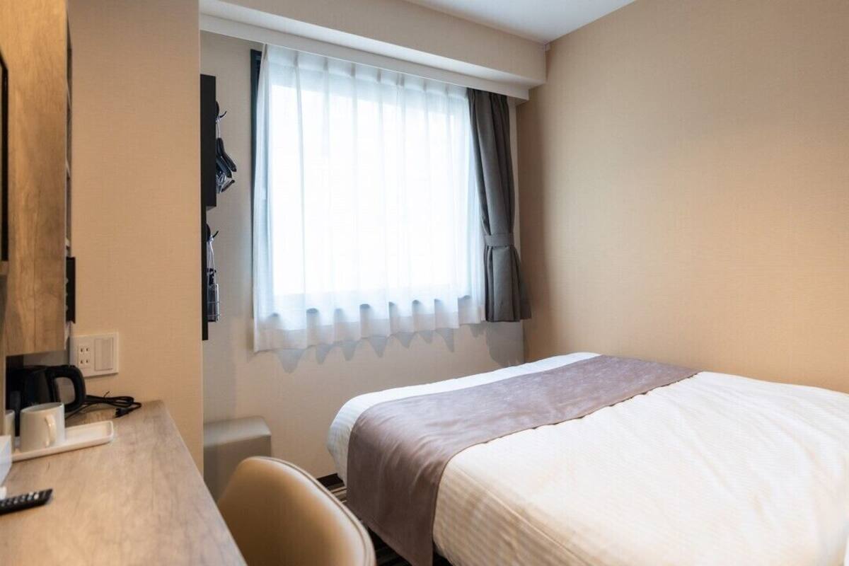 Quarto de hotel com cama, escrivaninha e cadeira, janela com cortinas e luminária na parede.